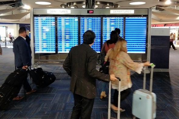 People standing in front of the arrivals departures boards in a busy airport checking their ...