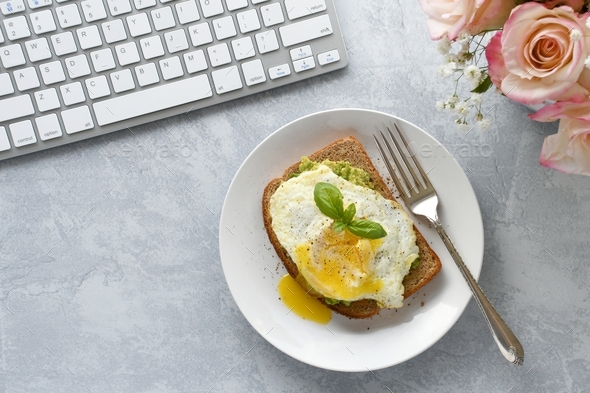 Avocado toast with fried egg over easy on desk with computer keyboard ...