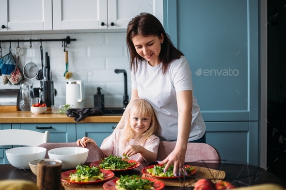A little blonde girl helps her mother prepare dinner in the kitchen ...