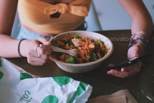 Young girl is eating take out or delivery food at home and using mobile ...