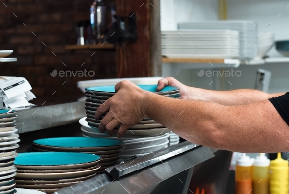 Restaurant kitchen employee placing stack of clean dishes on a shelf ...