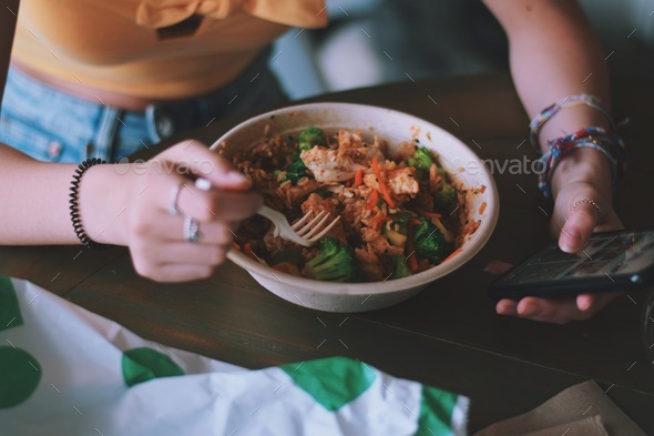 Young girl is eating take out or delivery food at home and using mobile ...