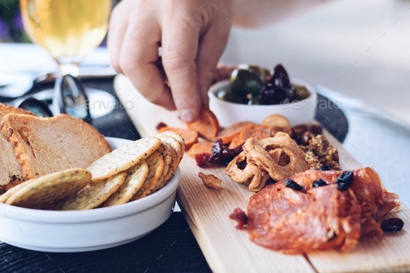 Person’s hand reaching for cured sausage on Charcuterie board with ...