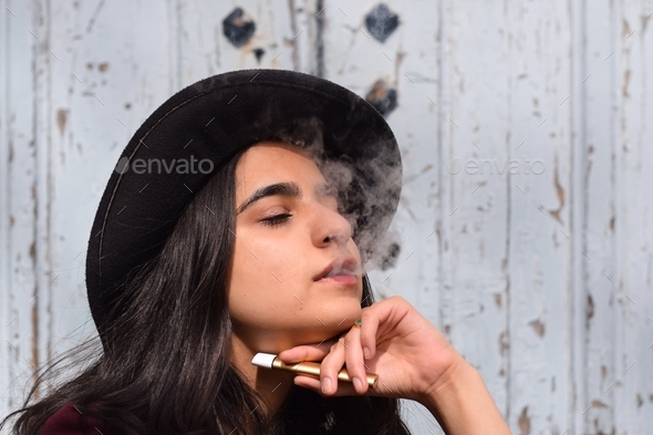 Closeup portrait of young girl in black fedora hat smoking vaping e ...