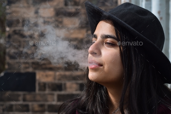 Closeup portrait of young girl in black fedora hat smoking or vaping ...