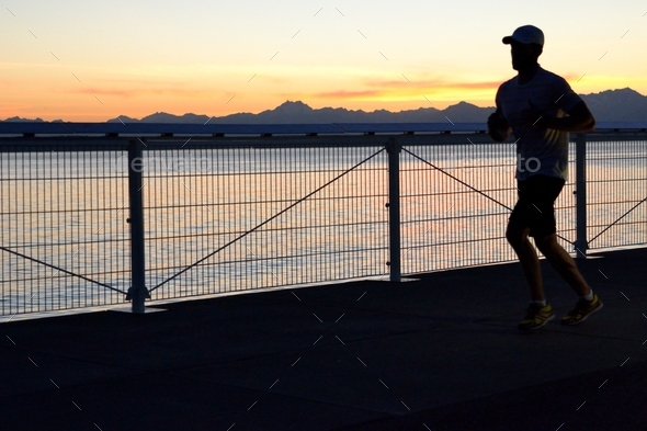 Silhouetted man wearing a baseball cap jogging in golden hour sunset ...