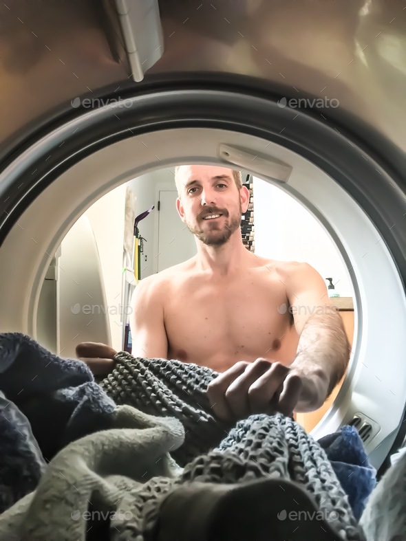Smiling shirtless man pulling laundry out of dryer. Stock Photo by RLTheis