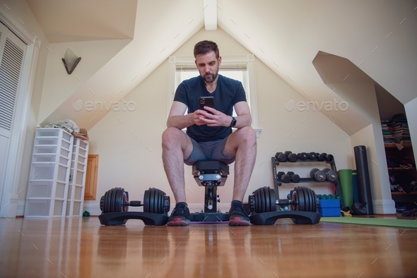 Man using smart phone while sitting on exercise bench in home gym with ...