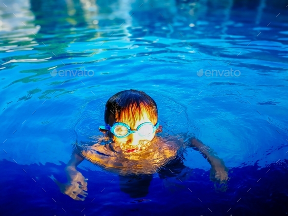 young boy swimming with goggles in a blue swimming pool with sunlight ...