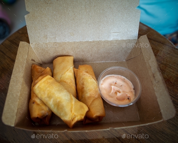 Fried spring rolls in a brown takeaway container with creamy sauce ...