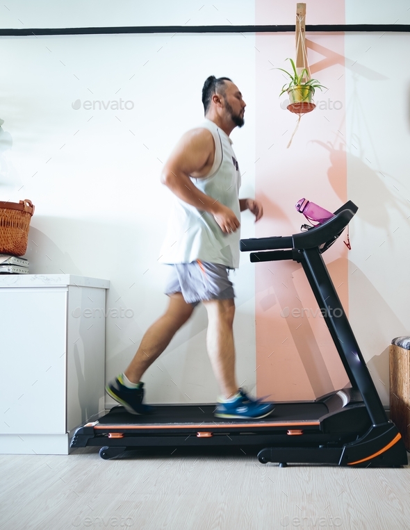 Full-length profile shot of a young man running on a treadmill at home ...