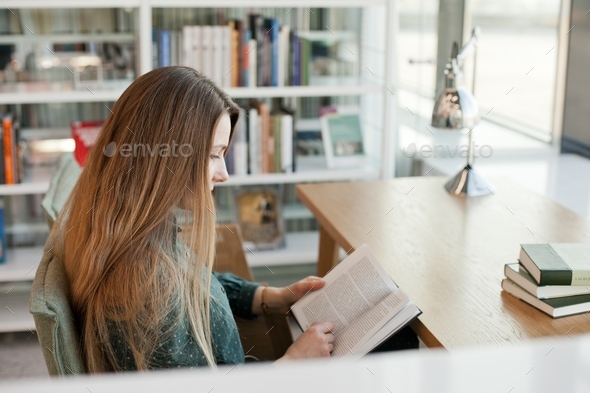 Student studying at a public library: reading a book at a desk with ...