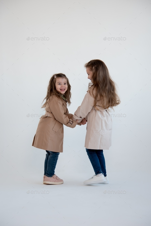 Two cute little girls in identical trench coats stand together. White ...