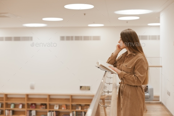 Young woman student in the public library standing with book by the ...