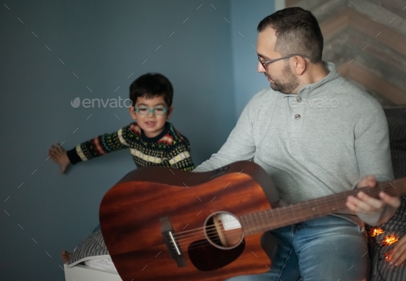 a father playing guitar to his son, father teaching son how to play a ...