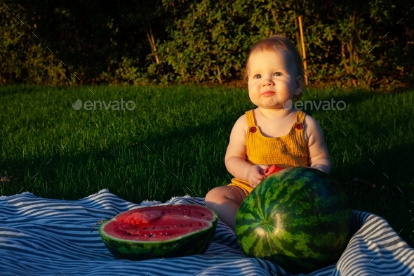little kid eats a piece of watermelon sitting next to a big watermelon ...