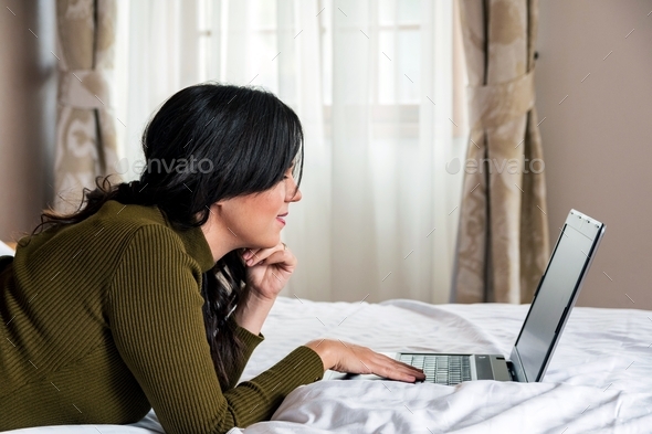 Side view of beautiful young woman laying on bed, using laptop computer ...