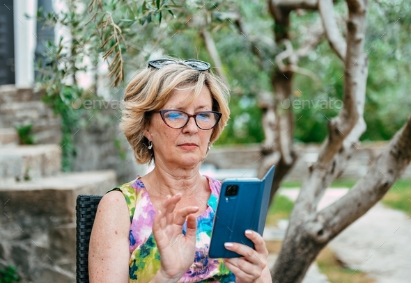 Senior woman sitting under olive tree in back yard, using mobile phone ...