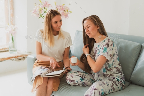 two adorable young women chatting at home on the sofa, drinking coffee ...
