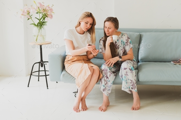 two adorable young women chatting at home on the couch, drinking coffee ...