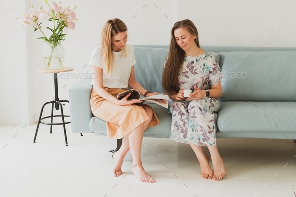 two adorable young women chatting at home on the sofa, drinking coffee ...