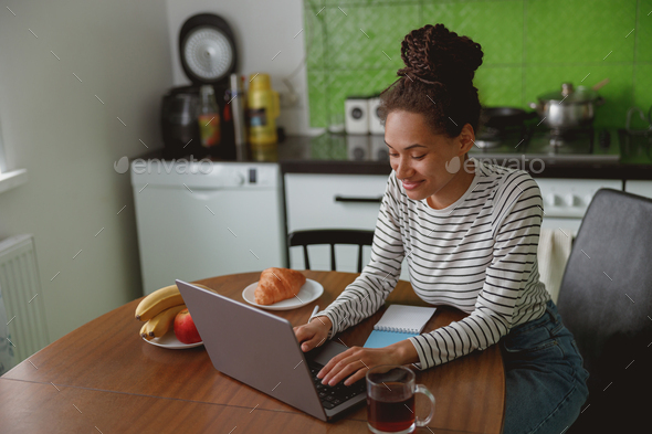 Young positive woman working on computer sitting at table in kitchen in ...