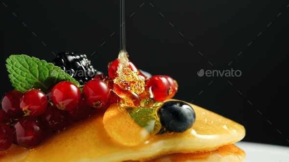 Stack of fresh fluffy pancakes decorated on top with forest berries and pouring honey syrup ...