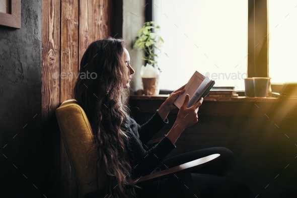 Happy young millennial woman with long curly hair reading book in a ...