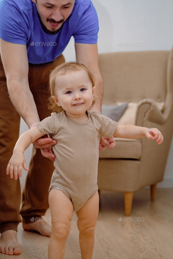 A cheerful smiling baby learning to walk supported by father. Father ...