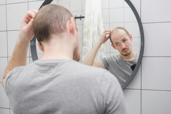 Stressed man with hair loss problem critically looking at mirror in the ...