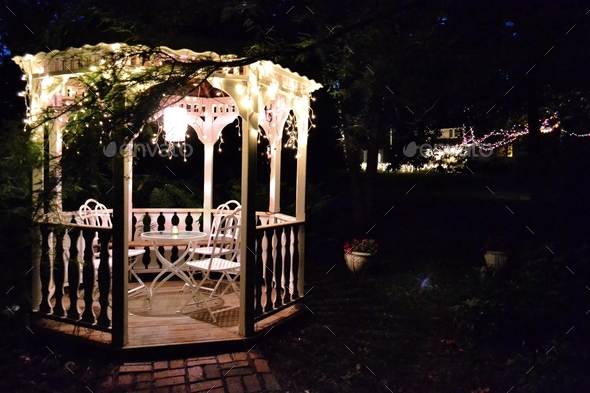 Gazebo in a garden at night with lights, a table and chairs with house ...