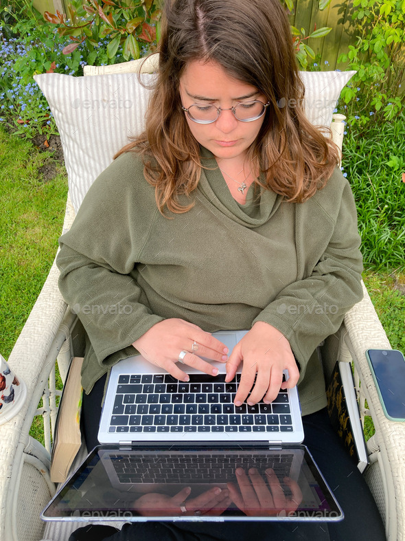 Gen Z woman sitting in garden using laptop computer with cellphone by ...