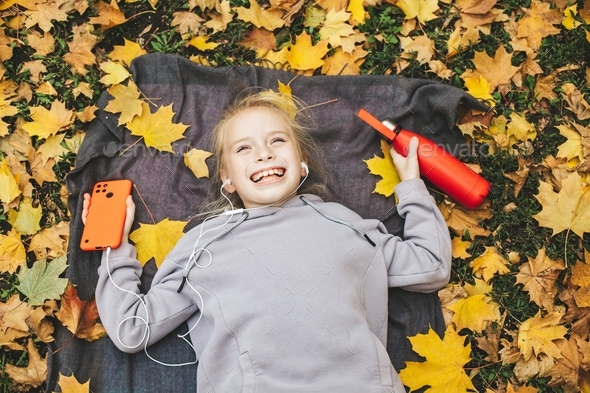 Happy blonde gen z girl lay on yellow autumn leaves with red bottle and ...