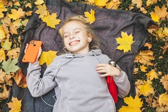 Happy blonde gen z girl lay on yellow autumn leaves with red bottle and ...