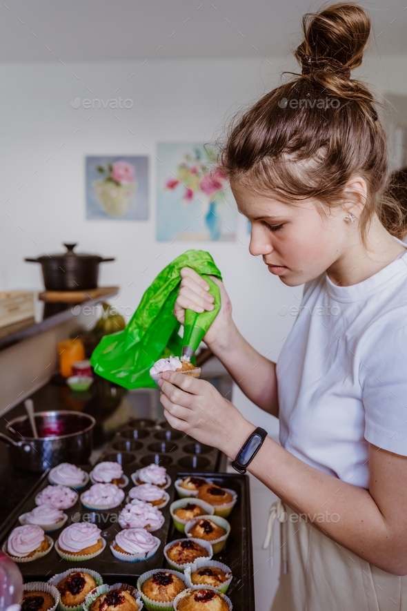 Tween girl putting cream topping on top of homemade cupcakes at kitchen ...