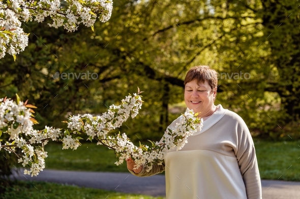Senior woman smelling white blooming tree in the park. Seasonal allergy ...