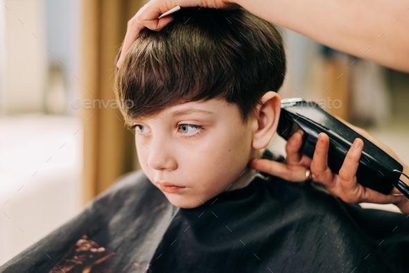 Little kid boy in the barbershop or hairdressing saloon. Barber is ...