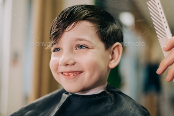 Little kid boy in the barbershop or hairdressing saloon. Barber is ...