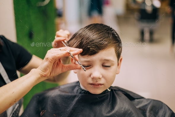 Little kid boy in the barbershop or hairdressing saloon. Barber is ...