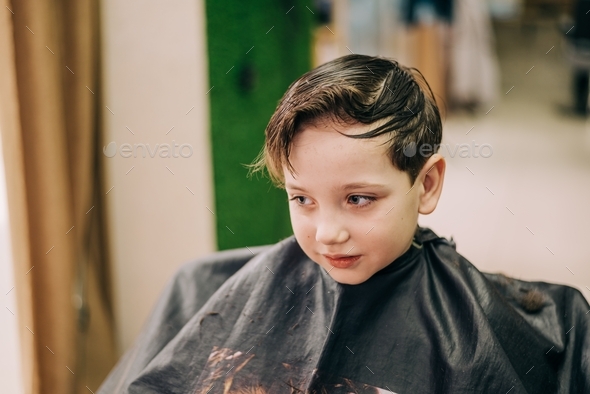 Little kid boy in the barbershop or hairdressing saloon. Barber is ...
