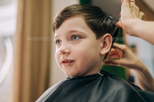 Little kid boy in the barbershop or hairdressing saloon. Barber is ...