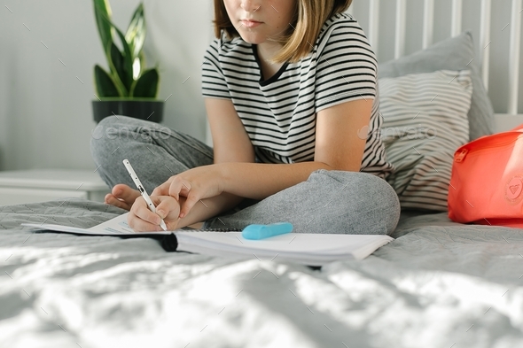 Focused schoolgirl in casual clothing doing homework, sitting on bed in ...