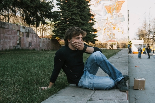 upset guy sits on the curb in the city and covers his mouth with his ...