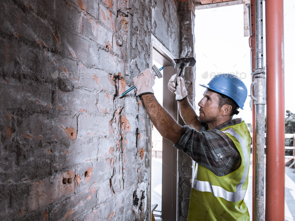 A worker opens a hole in a brick wall using a chisel and hammer in a ...
