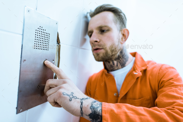 tattooed criminal in orange uniform pressing button in prison cell ...