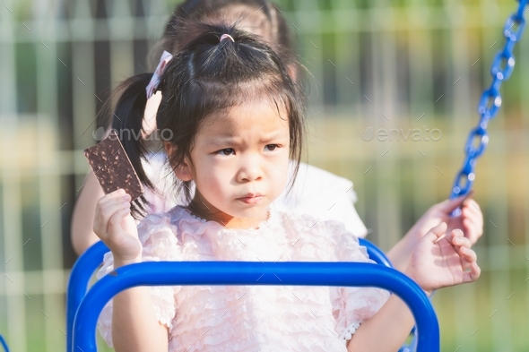Cute Asian girl is sitting on playground swing while eating chocolate ...