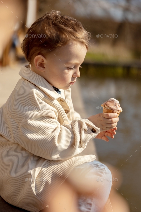 Little adorable boy sitting outdoors and eating ice cream. Lake, water ...