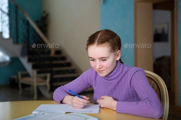 Teenage girl doing homework. She sits at the table at home and writes ...