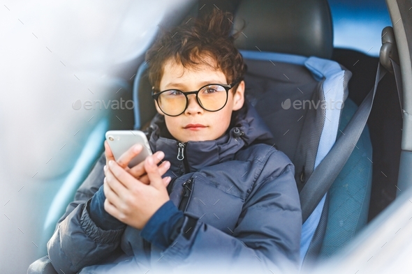 Curly boy with eyeglasses using mobile phone while sitting in blue car ...