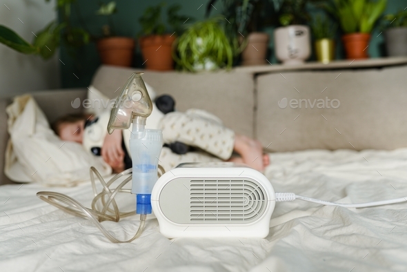 Sick boy laying in bed with his bear toy and nebulizer inhaler. Unwell ...